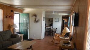 Uma sala de estar acolhedora com piso de madeira, uma porta verde, um espaço de jantar com mesa e cadeiras, e uma cozinha ao fundo.
