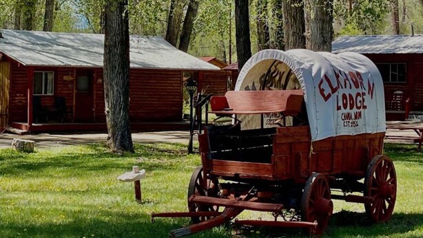 Uma carruagem coberta com uma placa que diz “ELK HORN LOAGE, CNY, NM EST. 1854” em uma área gramada, com cabanas de madeira ao fundo.