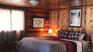Quarto estilo cabana de madeira com uma cama, mesa de cabeceira, abajur, janela e quadro na parede.