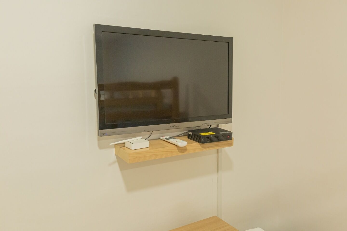 Televisão de tela plana fixada na parede, com uma prateleira de madeira embaixo, onde estão um aparelho de DVD e um controle remoto.