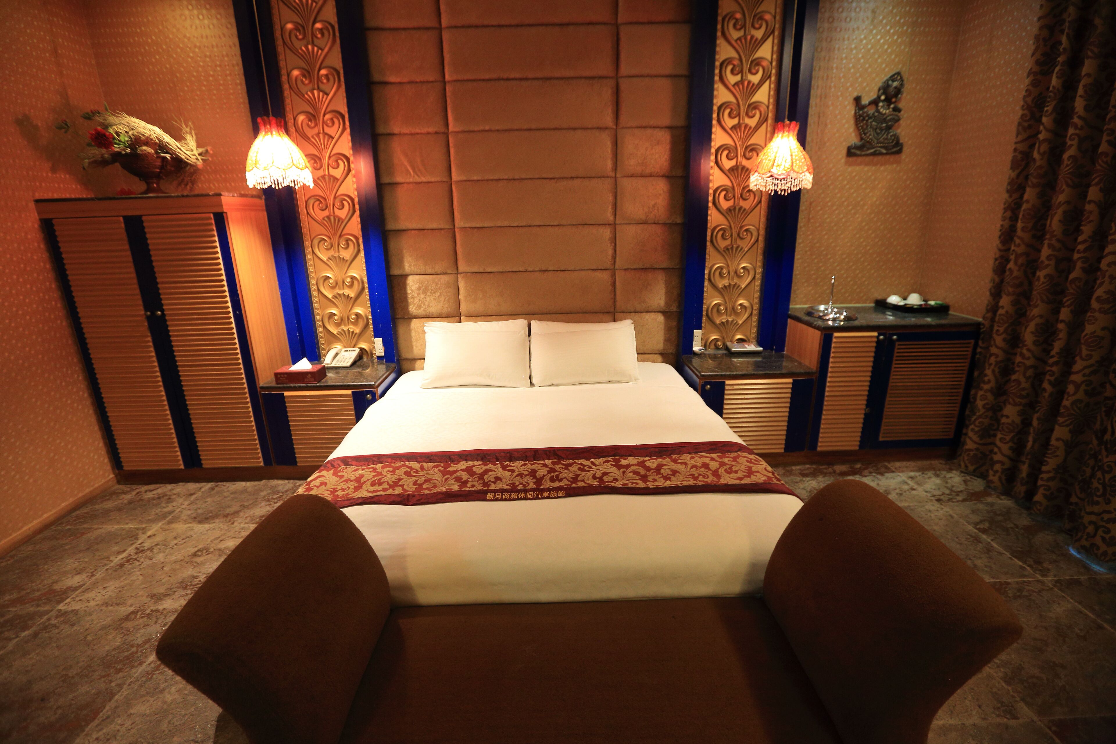 Quarto de hotel com uma cama grande, duas mesas de cabeceira, um criado-mudo, uma luminária decorativa e uma pequena mesa com abajur.