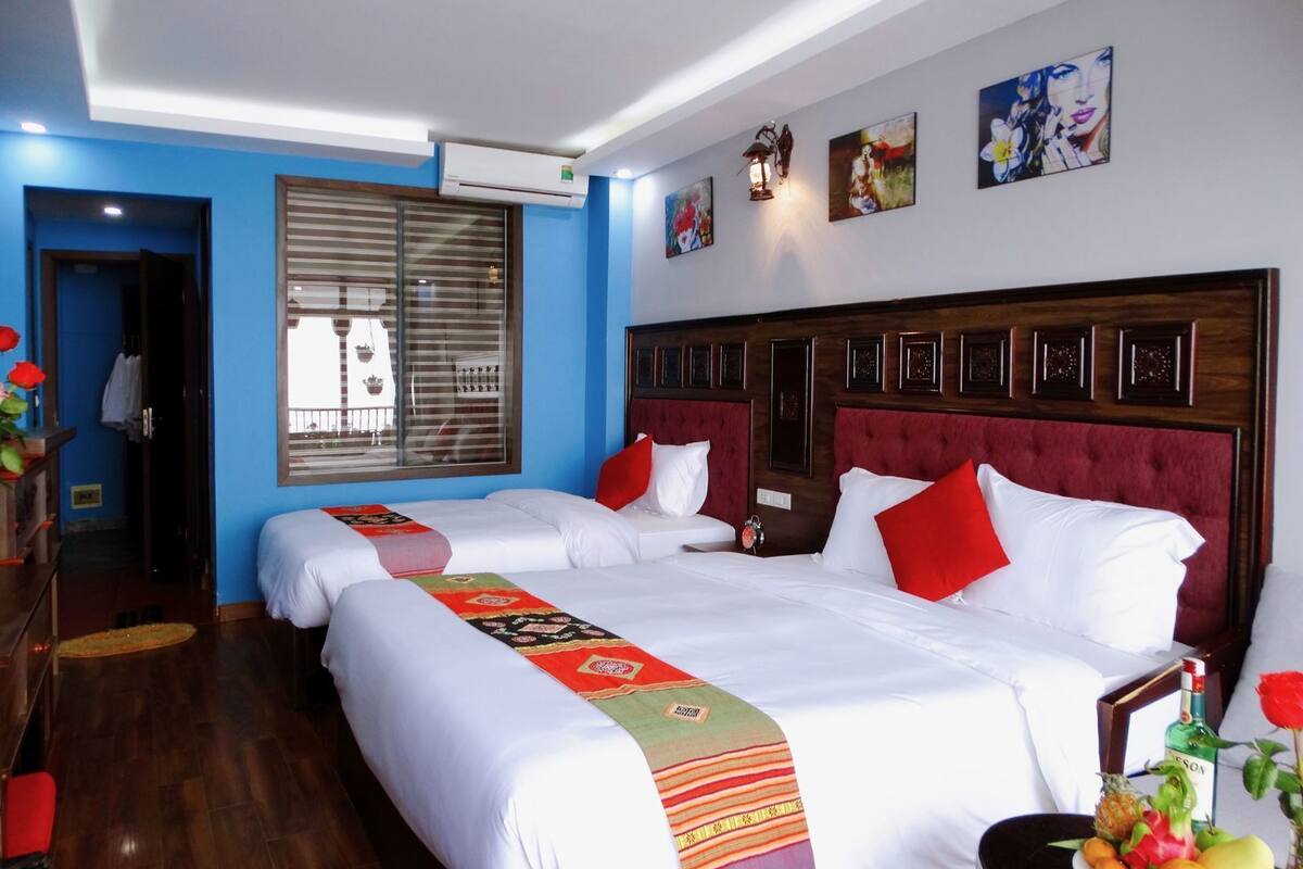 Quarto de hotel com duas camas, cabeceira de madeira, janela com persianas e obras de arte nas paredes.