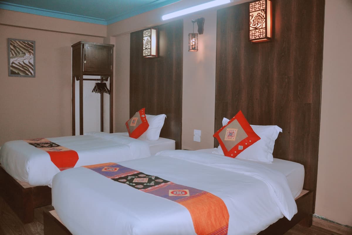Quarto de hotel com duas camas, cada uma com almofada decorativa e um cobertor. Há luminárias fixadas na parede e um armário de madeira.
