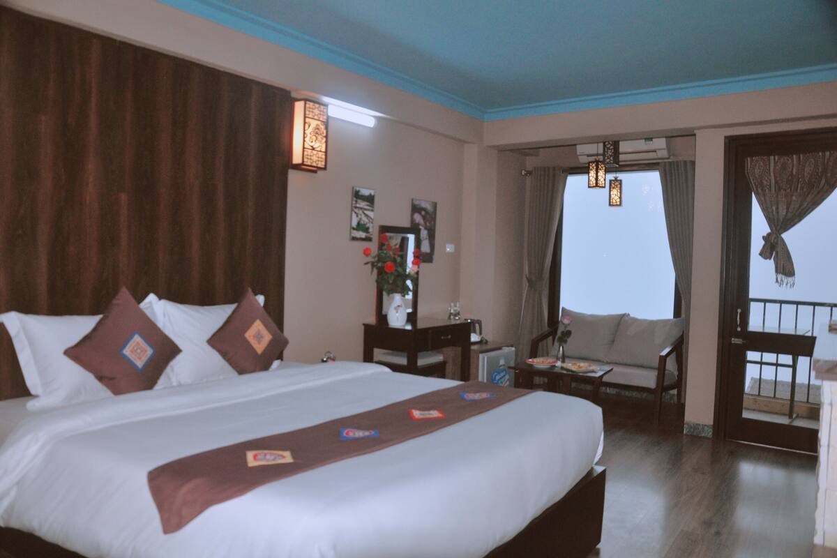 Quarto de hotel com uma cama grande, uma escrivaninha com cadeira, uma mesinha com cadeira e uma varanda com vista.