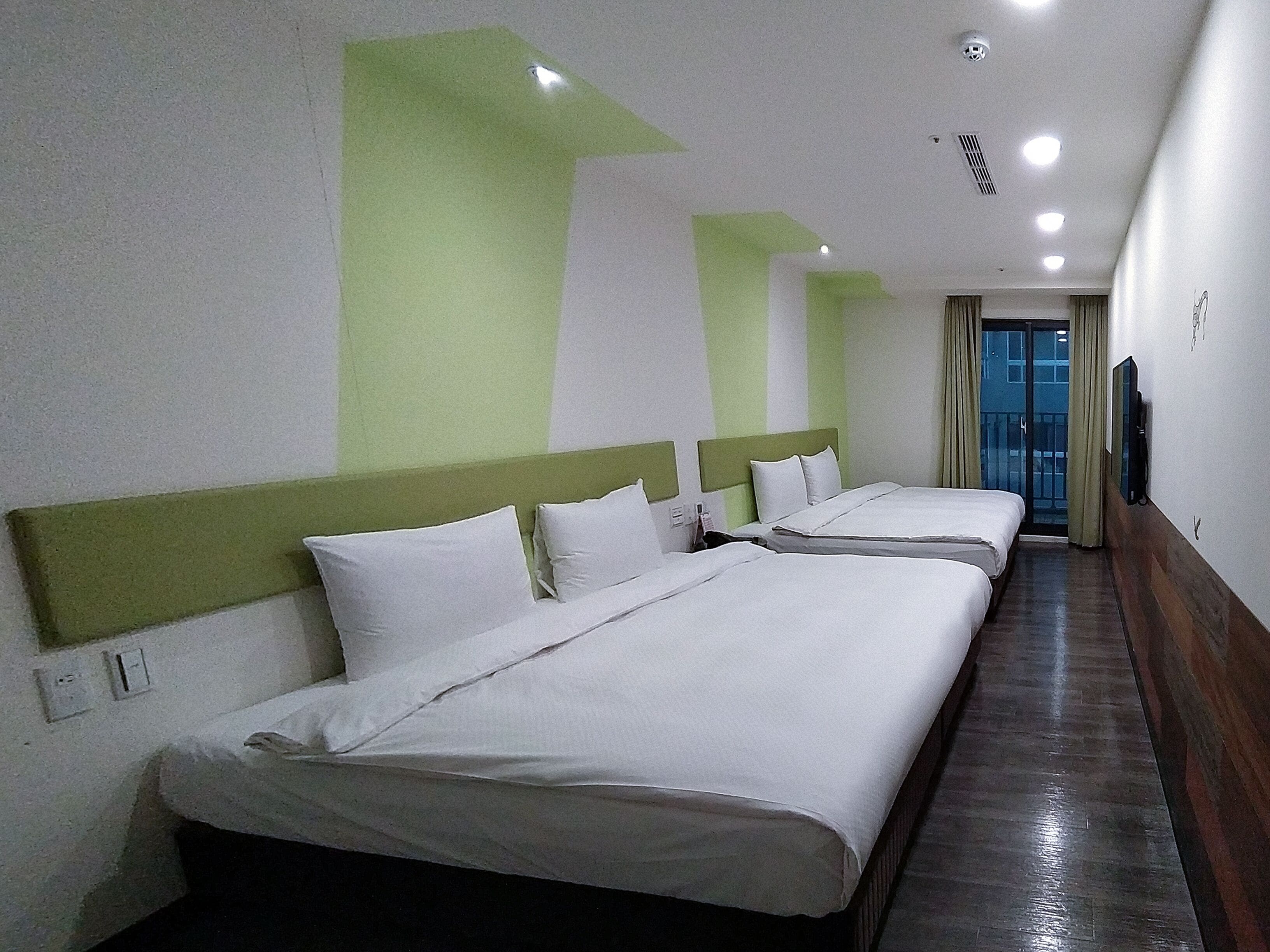 Quarto de hotel com três camas de solteiro, piso de madeira, uma parede de destaque verde e uma porta que dá para uma varanda.