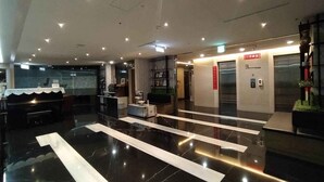Um hall moderno com balcão de recepção, totem informativo digital, área de espera e elevador.