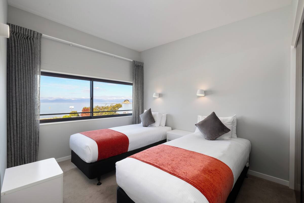 Quarto de hotel com duas camas, vista para o mar e uma escrivaninha branca.