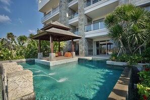 Uma área de piscina com um bangalô, espreguiçadeiras e um edifício moderno com sacadas.