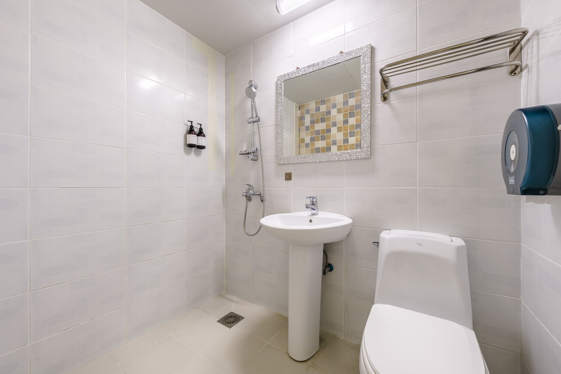Casa de banho com azulejos brancos, lavatório suspenso, sanita, chuveiro com chuveiro de mão e barra de toalhas.