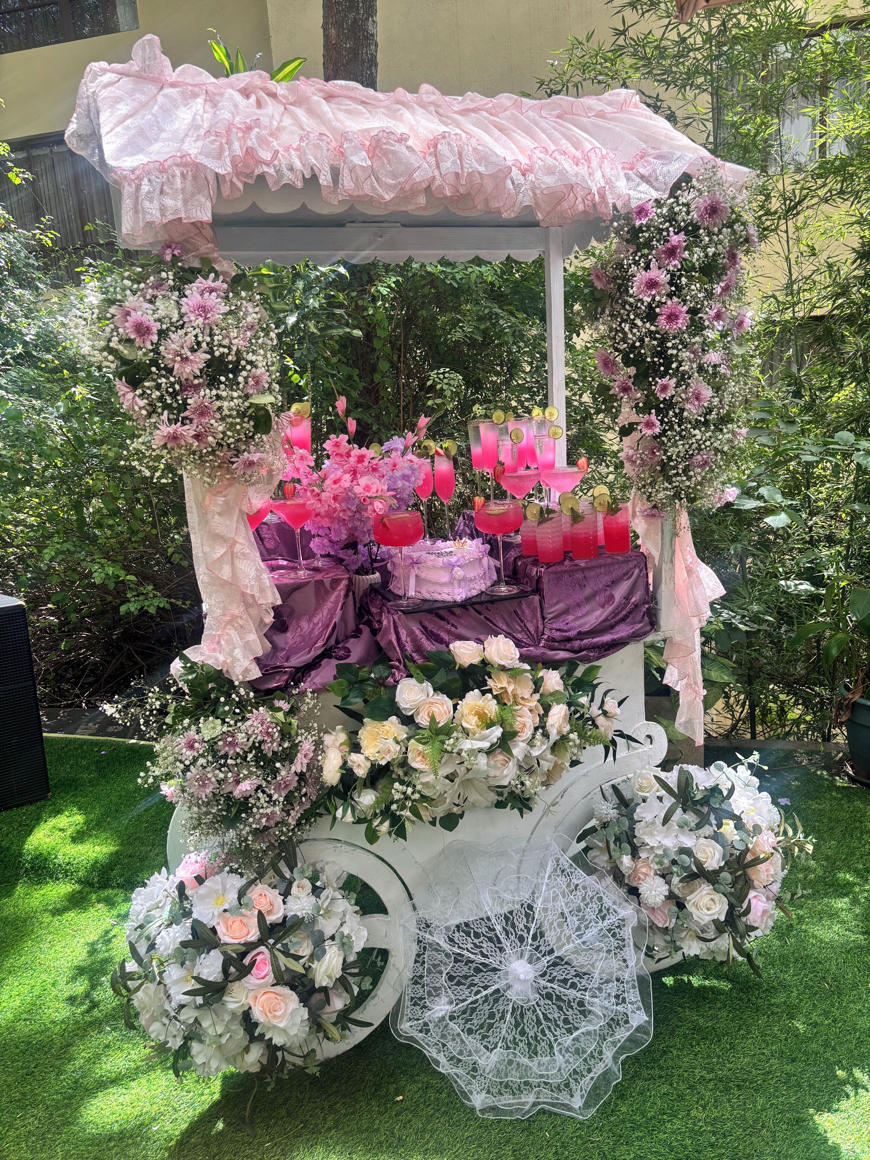 Um quiosque externo fixo com toldo branco, decorado com arranjos florais e bebidas cor-de-rosa.
