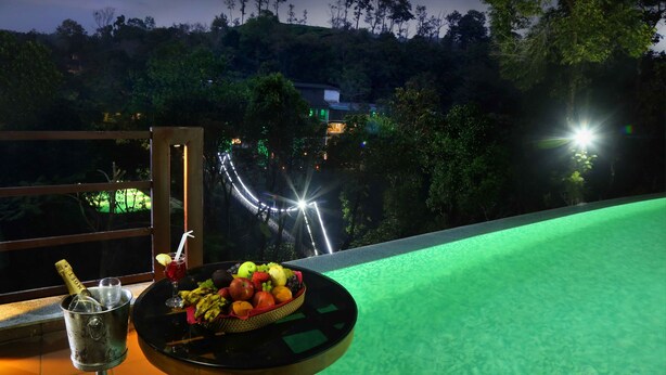 Uma mesa com uma fruteira, uma garrafa de champanhe e um copo de bebida vermelha, com vista para uma ponte iluminada e uma encosta arborizada.