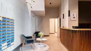 Uma área de recepção moderna com um balcão de madeira, uma cadeira azul, uma pequena mesa redonda com um vaso de flores e um aplique na parede.