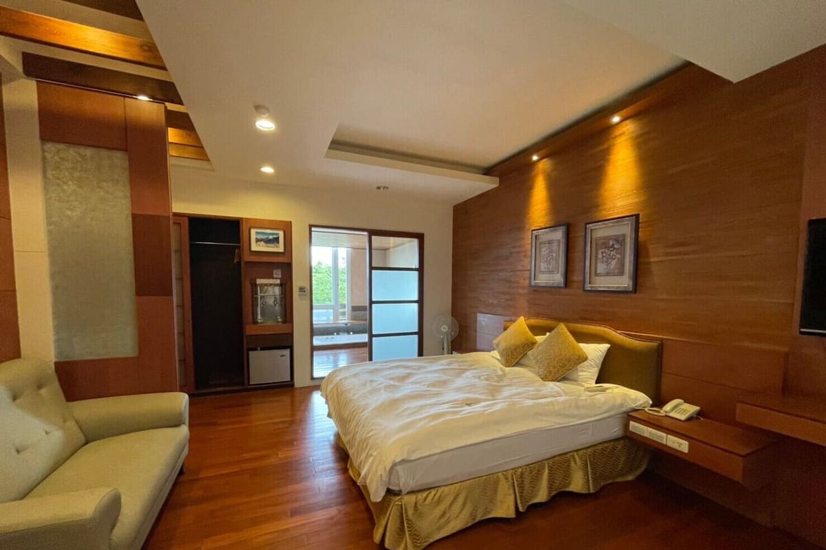Quarto com uma cama grande, um sofá, painéis de madeira e uma porta de vidro deslizante que dá para uma varanda.