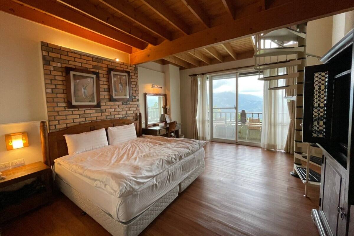 Quarto com cama grande, vigas de madeira no teto, parede de tijolos, varanda com vista e uma televisão.