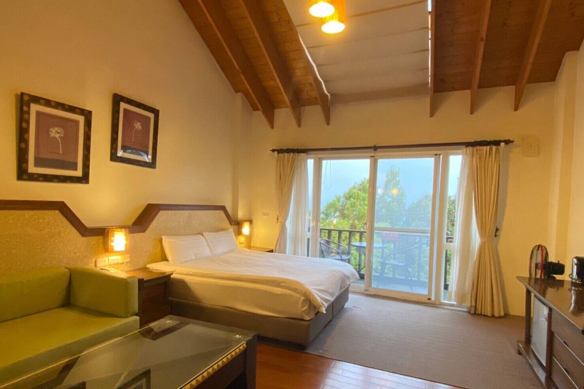 Quarto de hotel com cama, sofá verde, mesa de vidro e varanda com cortinas.
