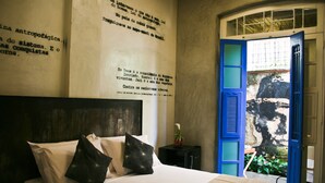Quarto com cama, almofadas, mesa de cabeceira e janela com persianas azuis.