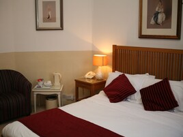 Quarto de hotel com uma cama, uma mesa de cabeceira com uma lâmpada, uma cadeira e uma pequena mesa com chaleira e telefone.