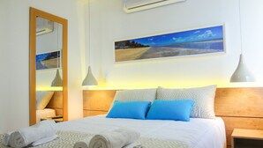 Cama bem arrumada com toalhas enroladas, cabeceira de madeira, ar condicionado fixado na parede, pintura emoldurada de paisagem e luminária pendente.