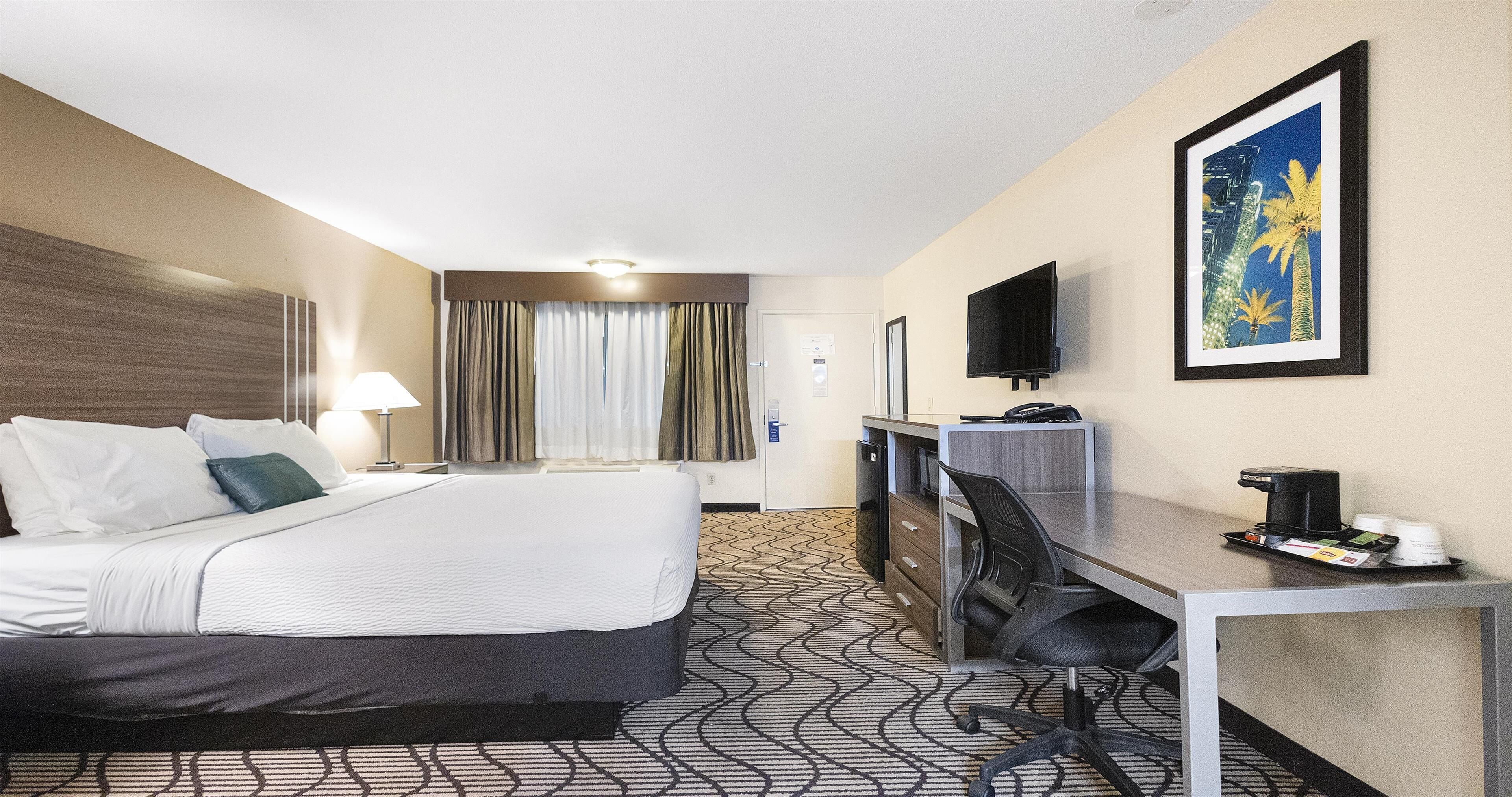 Quarto de hotel com cama, escrivaninha, cadeira, televisão e um quadro na parede.