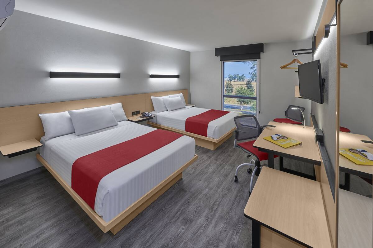 Quarto de hotel com duas camas, uma escrivaninha, uma cadeira, televisão e janela com vista.
