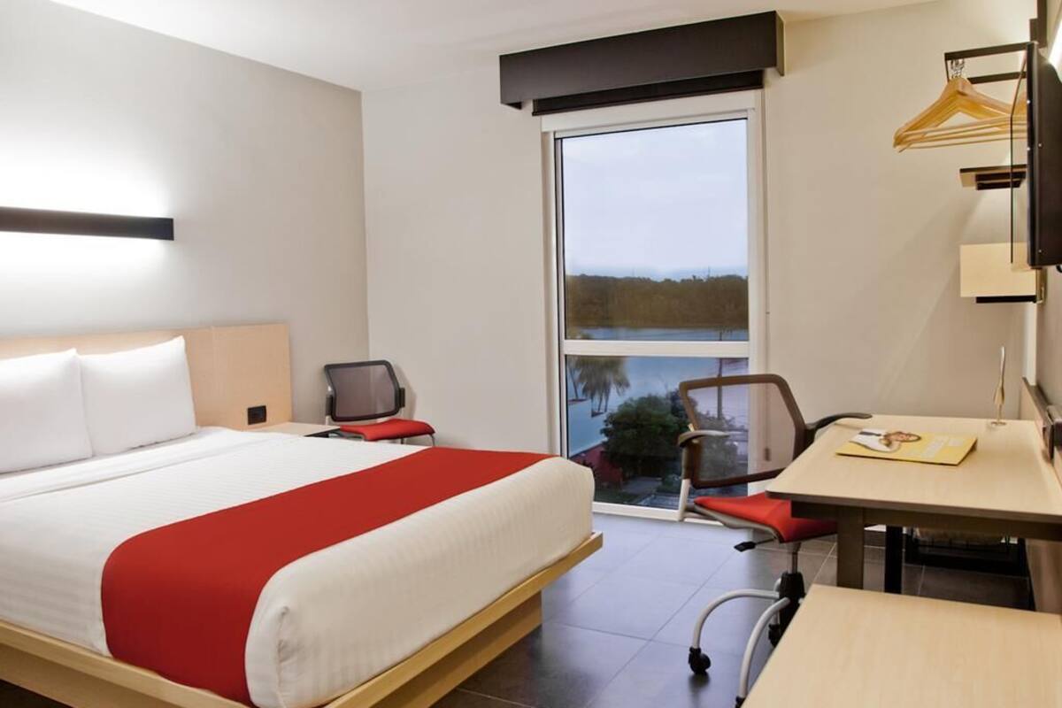 Quarto de hotel com cama, escrivaninha, cadeira, janela com vista e um cabideiro.