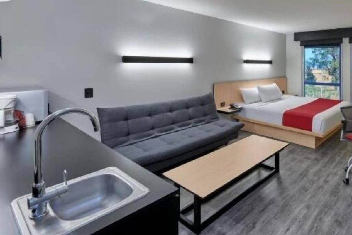Quarto de hotel compacto com cozinha compacta, um sofá, uma cama com manta vermelha, uma mesa com cadeira e vista para o exterior.