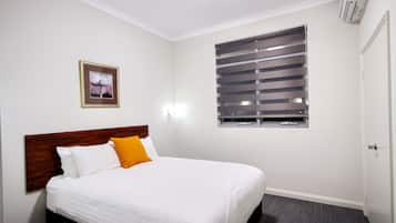 Uma cama bem arrumada com roupa de cama branca e laranja, um quadro na parede e uma janela com persianas.