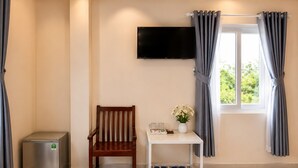 Quarto de hotel com duas camas, televisão, uma pequena mesa com um vaso de flores, uma cadeira, prateleira com sapatos e janela com cortinas.