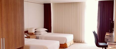 Quarto de hotel com duas camas, uma mesa, uma cadeira, minibar e uma janela grande com cortinas.