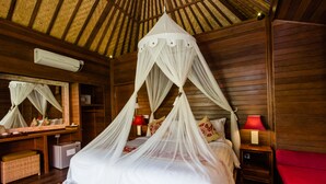 Quarto de madeira com uma cama de dossel, um espelho, uma mesa de cabeceira com abajur e um banco vermelho.