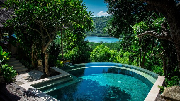 Piscina azul translúcida rodeada por vegetação exuberante e um deck de madeira.
