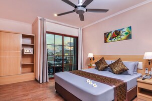 Quarto de hotel com cama, ventilador de teto, pintura, mesa de cabeceira e vista através de uma porta de vidro.