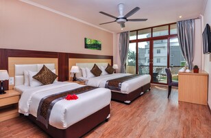 Quarto de hotel com duas camas, um ventilador de teto, uma janela grande com cortinas, uma mesa de trabalho e uma televisão.