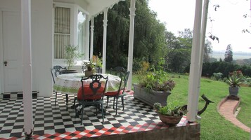 Alpendre coberto com piso xadrez, mesa com cadeiras e plantas em vasos.