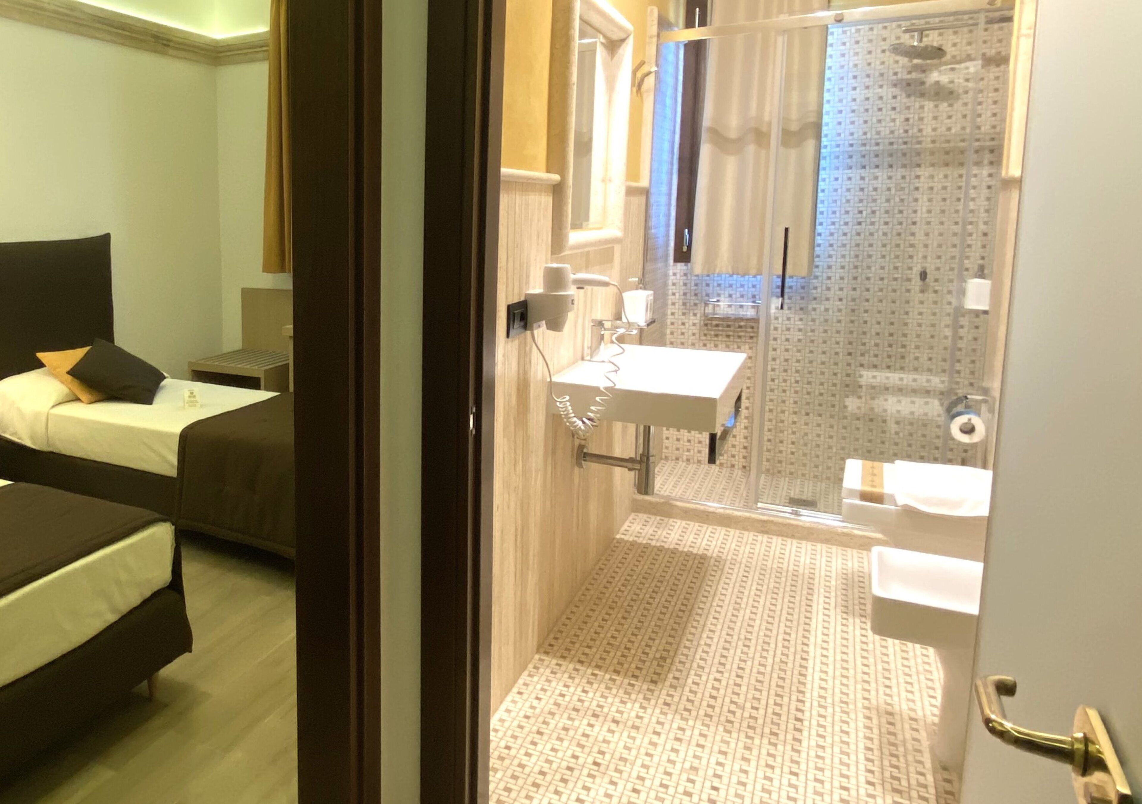 Quarto de hotel com cama, mesa de cabeceira e área de chuveiro com porta de vidro.