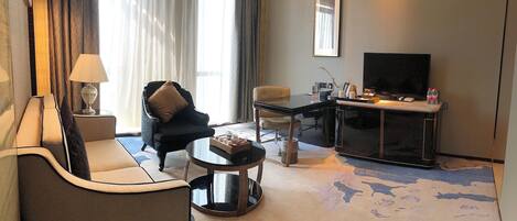 Quarto de hotel com sofá, poltrona, mesa de centro, escrivaninha e televisão.