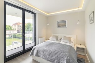 Quarto moderno com uma janela grande, cama com colcha cinza, duas mesas de cabeceira e uma pequena planta.
