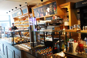Balcão de café com um quadro de cardápio, máquinas de café e diversos produtos de panificação.