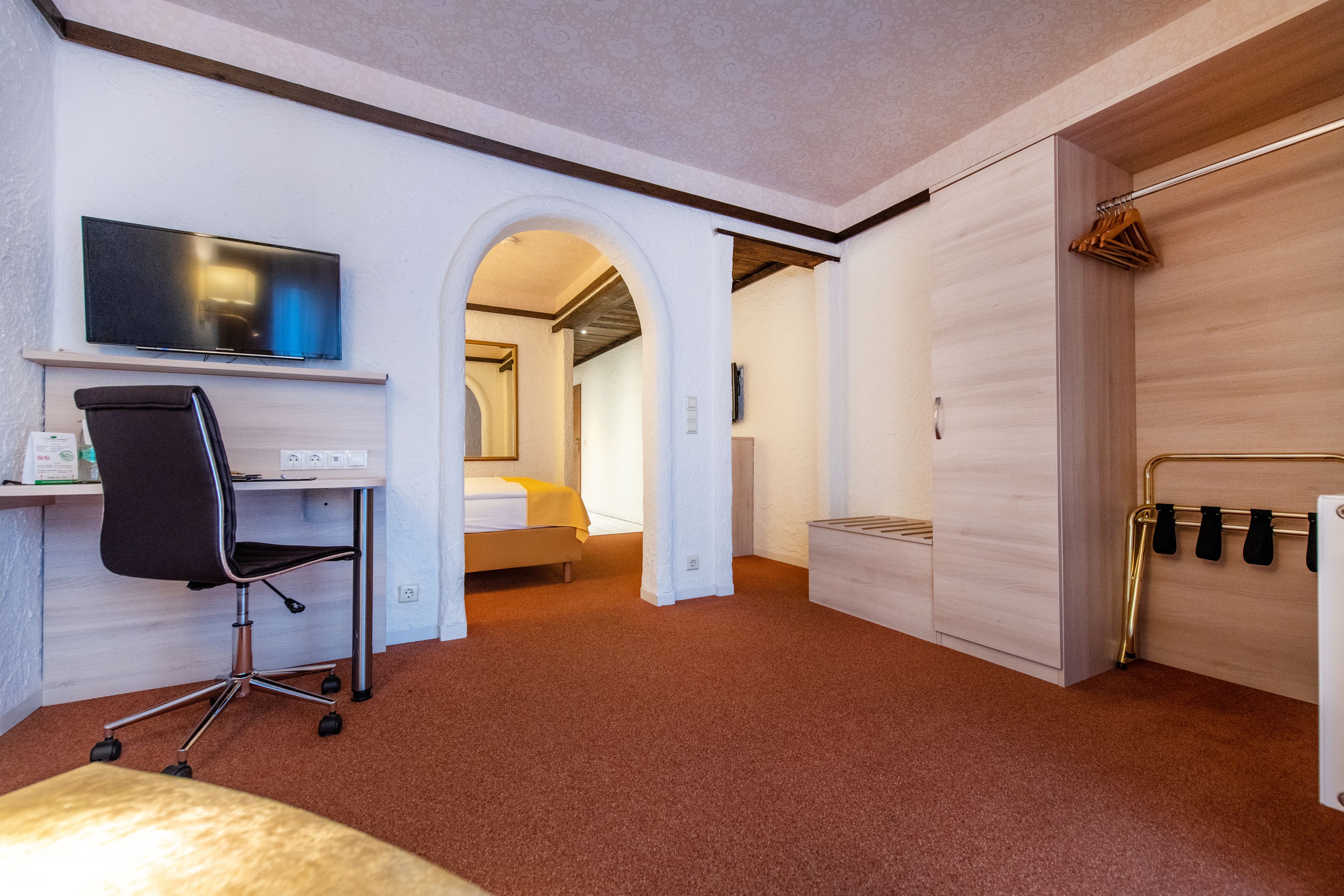 Quarto de hotel com secretária, cadeira, televisão, cama e guarda-roupa.