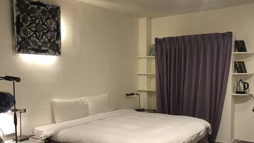 Cama bem arrumada com edredom branco e saia marrom, mesa de cabeceira com abajur, um objeto decorativo na parede, prateleira com livros e chaleira, e cortina escura cobrindo uma janela.
