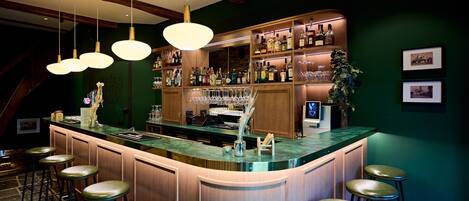 Uma área de bar com balcão curvo, vários taburetes, luminárias pendentes e prateleiras com garrafas e copos.