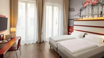 Quarto de hotel com duas camas, uma escrivaninha com computador, uma cadeira e uma janela grande com cortinas.