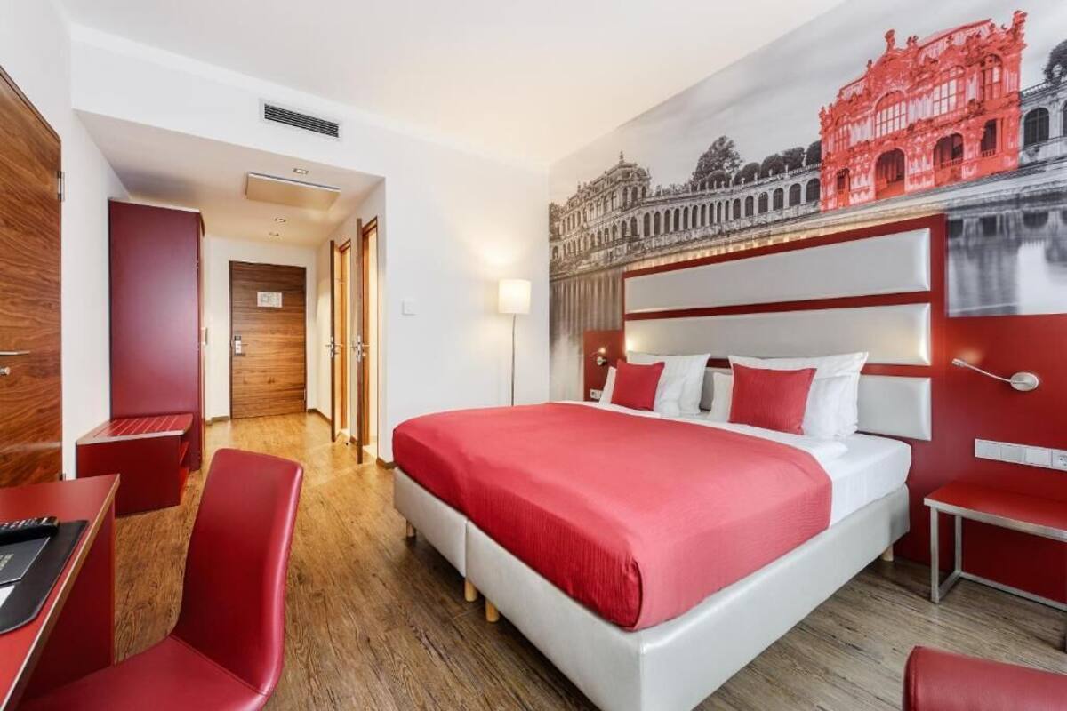 Quarto de hotel com uma cama grande, uma mesa e uma cadeira. Há uma porta que dá para outro cômodo.