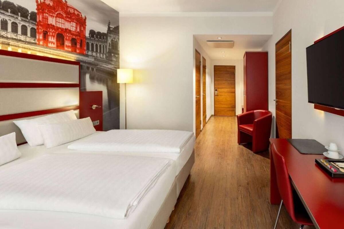 Quarto de hotel com duas camas, uma escrivaninha, uma cadeira, uma TV e um mural de um edifício.