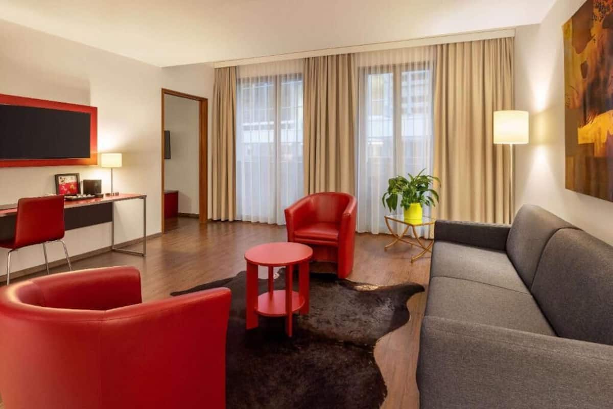Quarto de hotel moderno com sofá, cadeiras vermelhas, uma mesa pequena, escrivaninha e uma janela grande com cortinas.