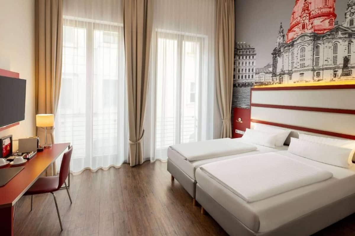 Quarto de hotel com duas camas, uma escrivaninha, uma cadeira e uma janela grande com cortinas.