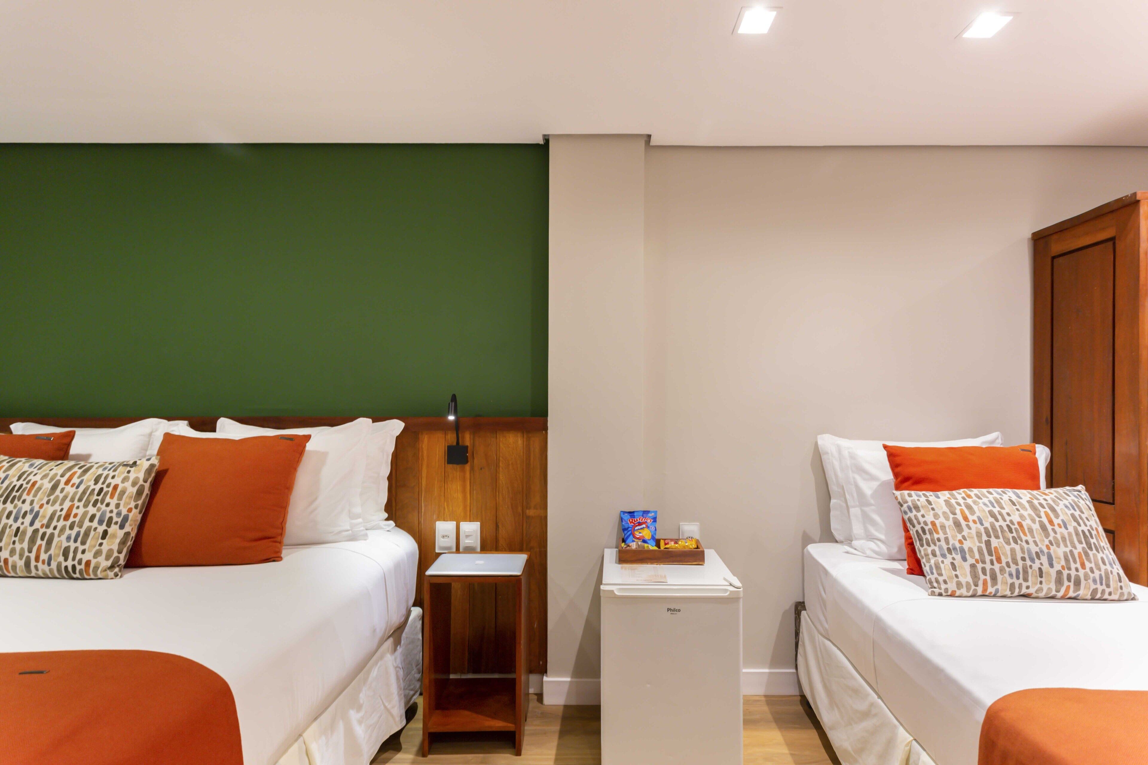 Quarto de hotel com duas camas, cada uma com roupa de cama branca e detalhes laranja. Há uma pequena mesa branca entre as camas e uma mesa de cabeceira de madeira com uma luminária.