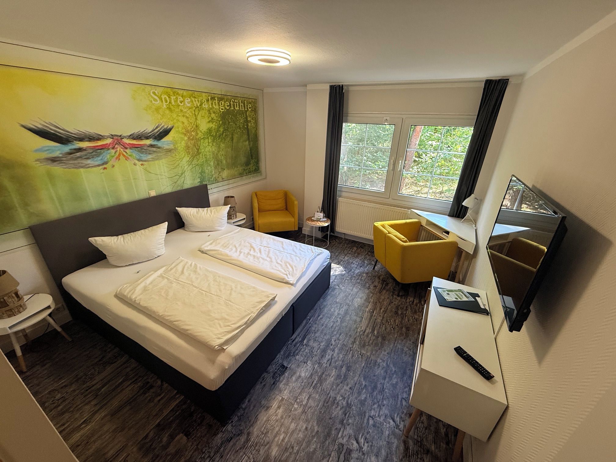 Quarto de hotel com uma cama grande, uma escrivaninha com cadeira, uma televisão e janela com vista para árvores.
