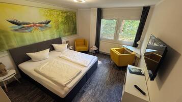 Quarto de hotel com uma cama grande, uma escrivaninha com cadeira, uma televisão e janela com vista para árvores.