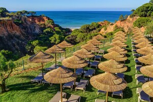 Um resort à beira-mar com fileiras de espreguiçadeiras e toldos de palha, com vista para um penhasco costeiro e o mar.